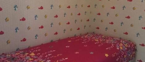 Quarto pequeno com uma cama de solteiro, papel de parede estampado com flores e colcha vermelha.