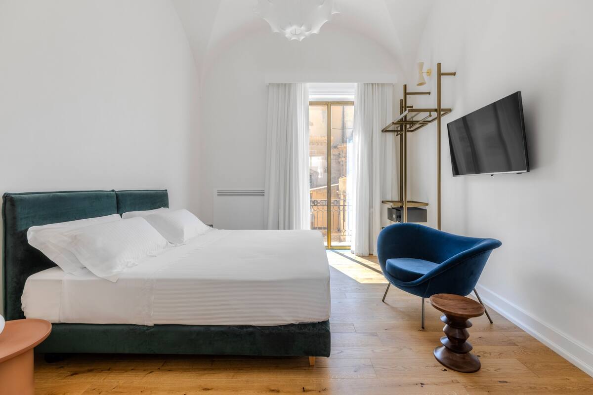 Quarto com cama, uma cadeira azul, televisão e piso de madeira.