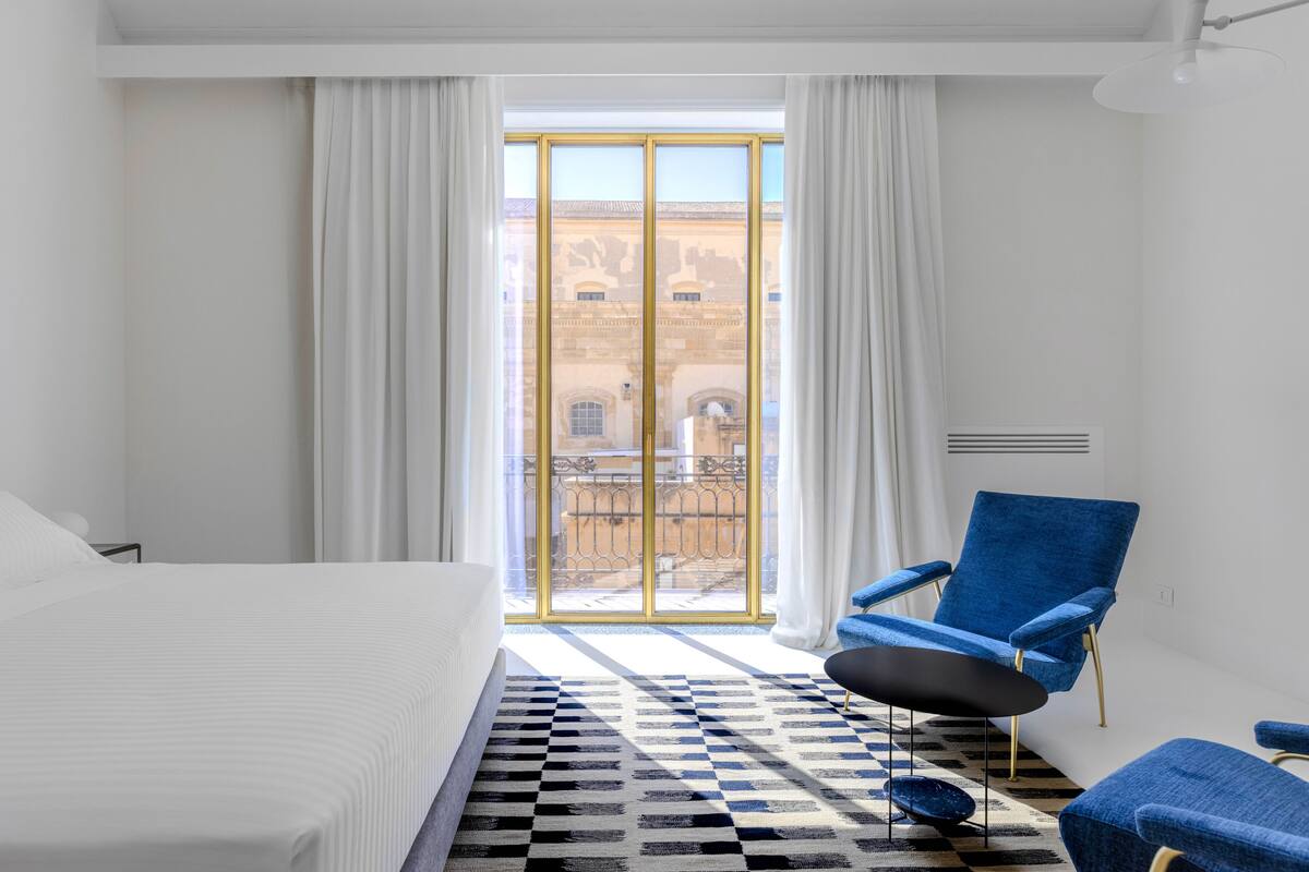 Quarto de hotel com cama, poltrona azul, tapete xadrez e vista para um edifício através de uma porta de vidro.