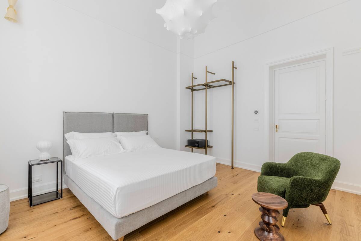 Quarto com cama, mesa de cabeceira, abajur, poltrona verde e porta branca.