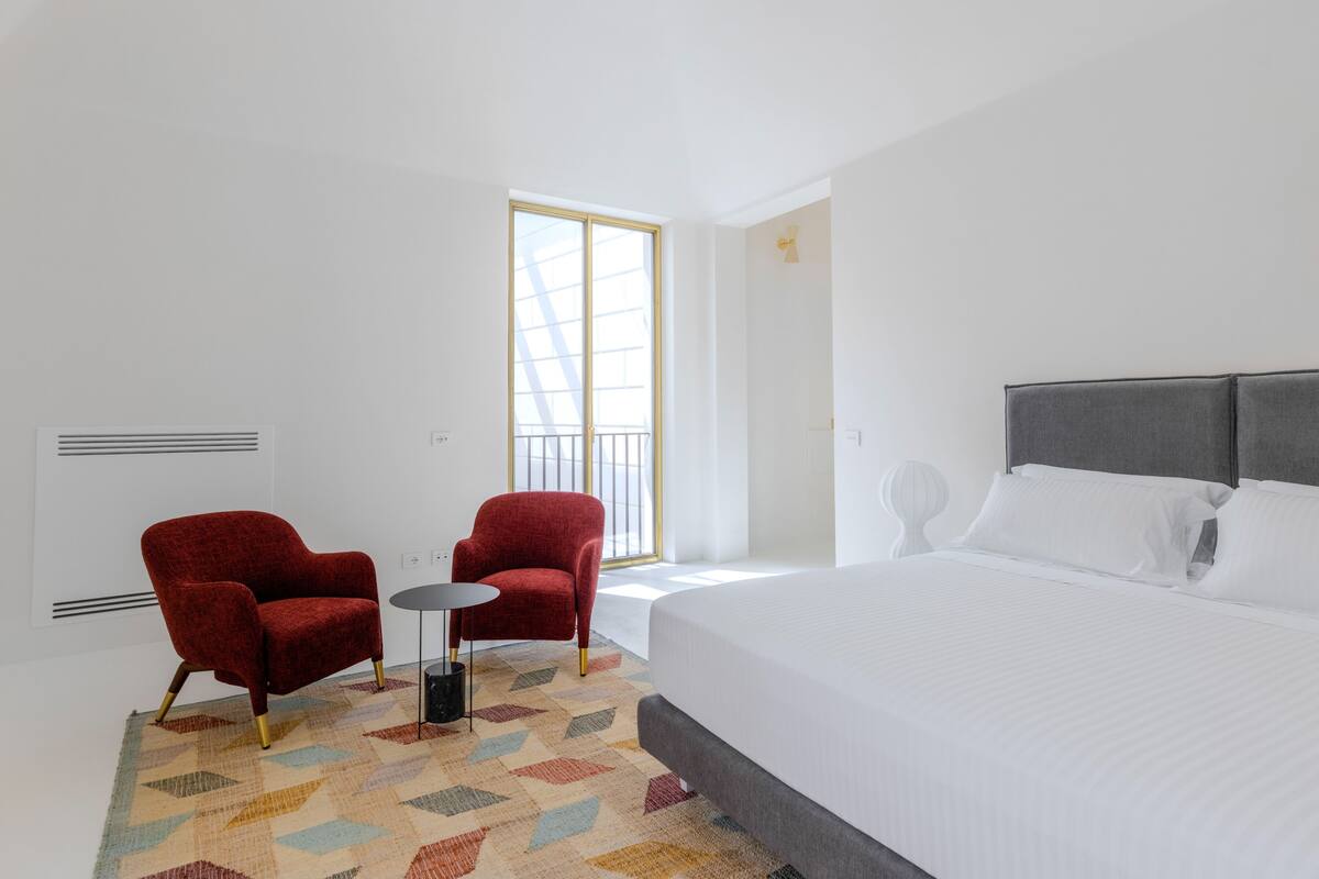 Quarto de hotel com uma cama grande, duas poltronas vermelhas, uma mesa redonda pequena e um tapete com padrão geométrico.