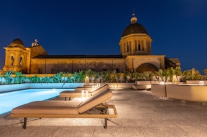 Uma área na piscina com espreguiçadeiras e um edifício imponente com arquitetura em cúpulas ao fundo.