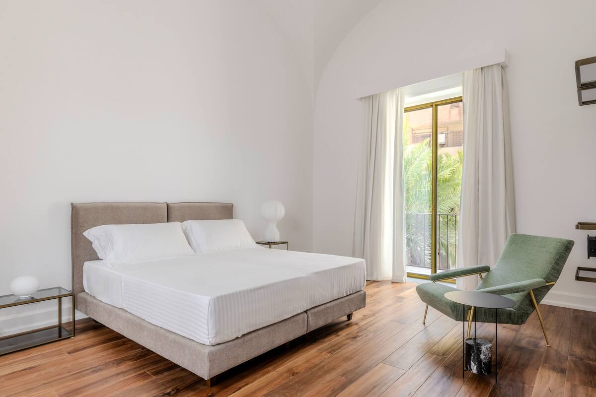 Quarto moderno com uma cama grande, uma poltrona verde e uma varanda com cortinas.