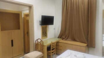 Quarto de hotel com escrivaninha, cadeira, televisão, guarda-roupa e janela com cortinas.