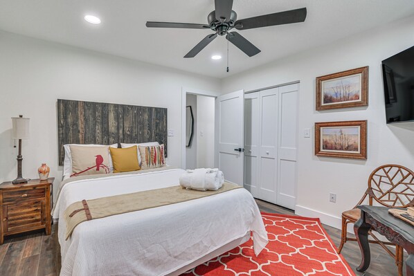 Quarto com cama, ventoinha no teto, mesa de cabeceira, luminária, tapete vermelho e uma cadeira.
