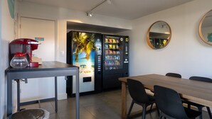 Um quarto com uma mesa de madeira, cadeiras pretas, uma máquina de café e máquinas de venda automática.