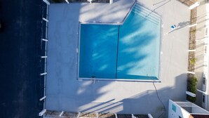 Vista aérea de uma piscina retangular com um deck ao redor, adjacente a um edifício com fachada branca e uma pequena área ajardinada.