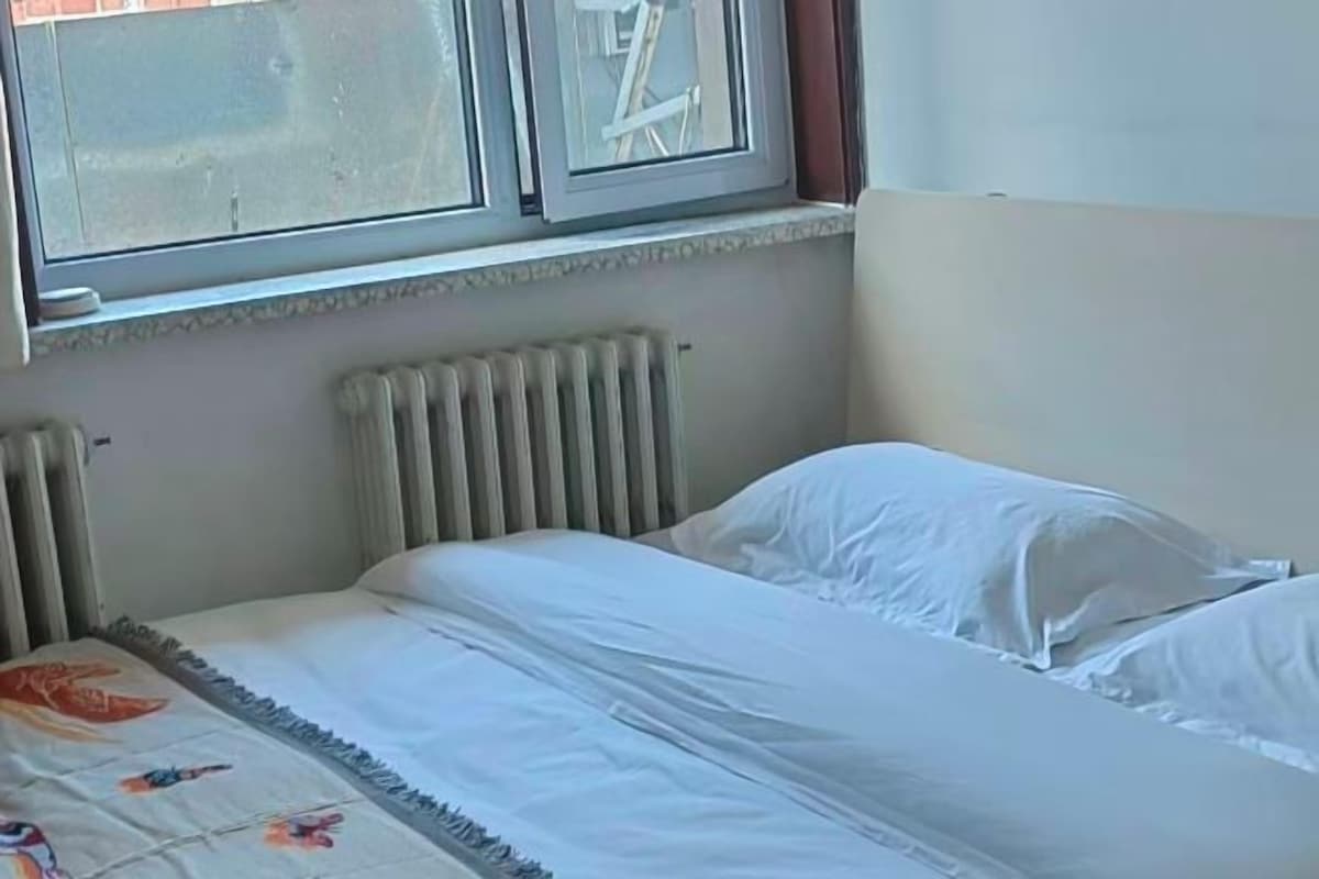 Uma cama de solteiro com edredom e almofadas brancas, um radiador embaixo e uma janela acima.