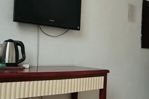 Televisão fixada na parede, uma mesa de madeira com uma chaleira e um pequeno objeto branco sobre a mesa.