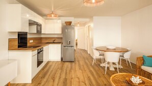 Cozinha moderna com armários brancos, bancada de madeira e forno embutido. Há uma área de jantar com mesa redonda e cadeiras brancas.