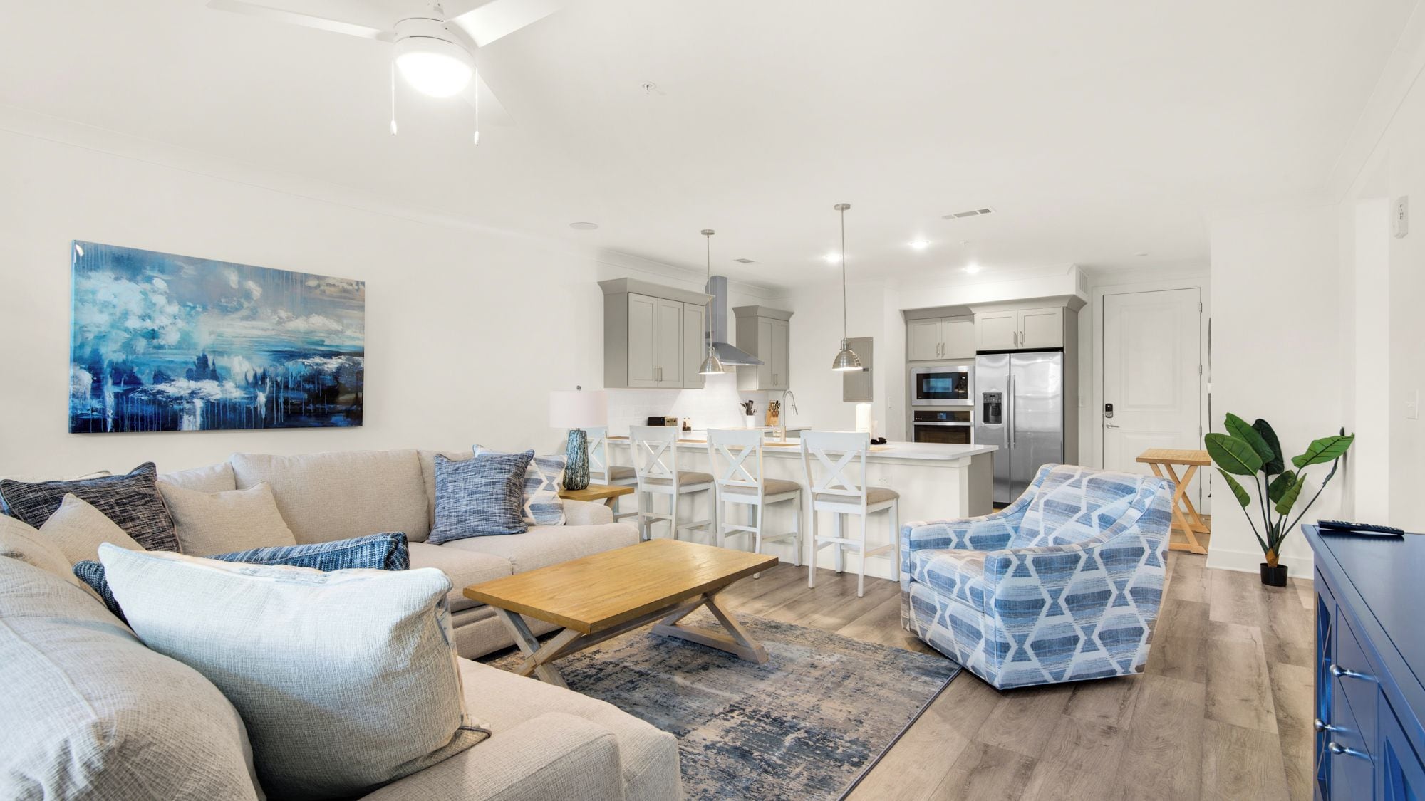 Uma sala de estar moderna com sofá, mesa de centro e poltrona estampada. Uma cozinha com armários e eletrodomésticos brancos é visível ao fundo.