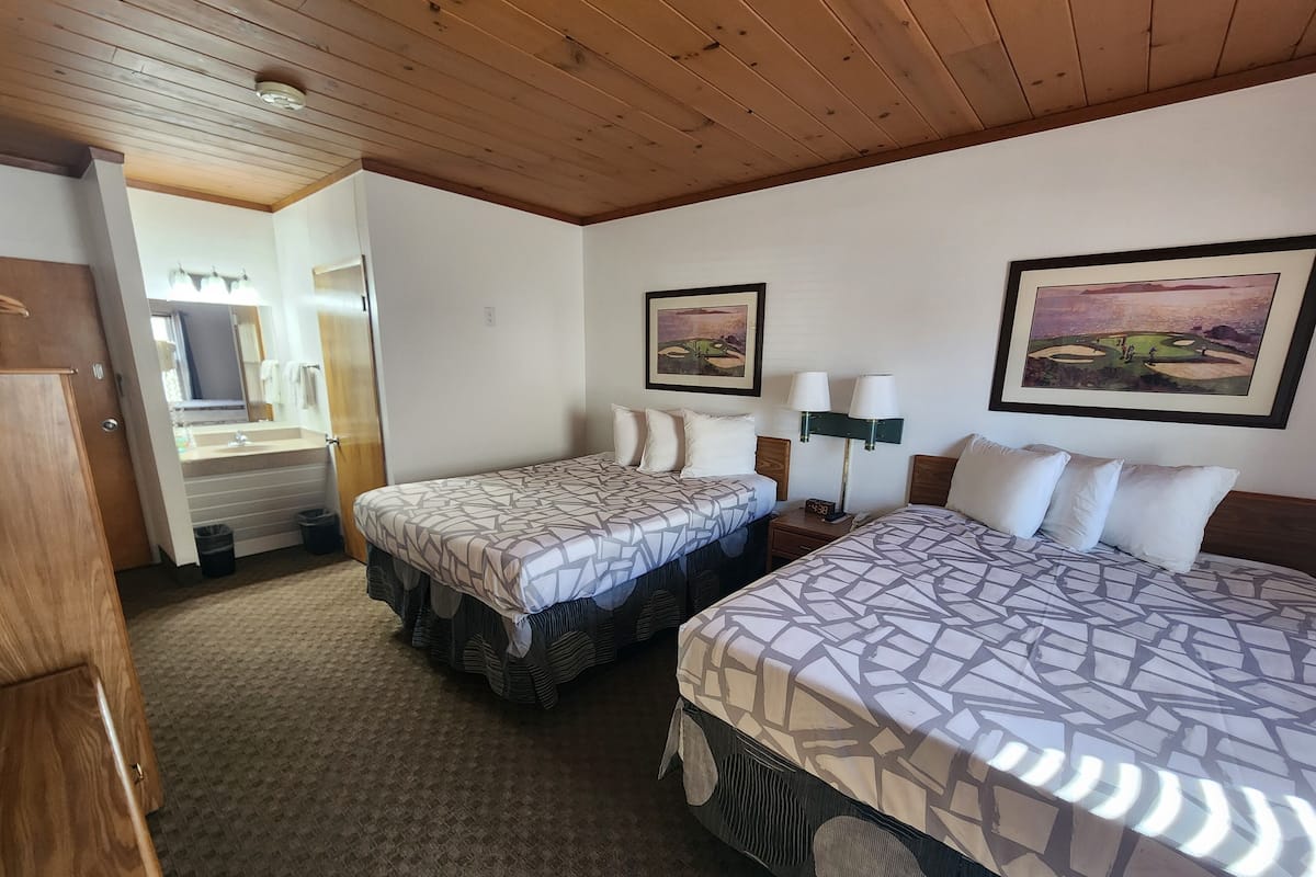 Quarto de hotel com duas camas, banheiro com pia e espelho, e um quadro na parede.