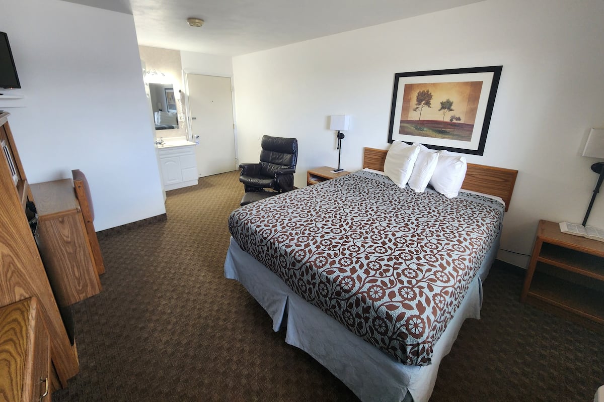Quarto de hotel com cama, uma cadeira, escrivaninha, espelho e um quadro na parede.