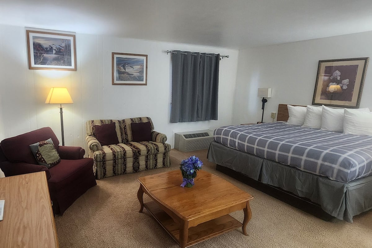 Quarto de hotel com cama, duas poltronas, uma mesa pequena e uma escrivaninha. Há luminárias, um espelho e quadros nas paredes.