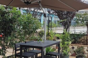 Um pátio externo com mesa e cadeiras, rodeado por plantas em vasos e vista para um edifício ao fundo.