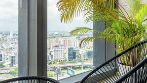 Varanda com duas cadeiras de vime preto, uma planta em vaso e vista para a cidade através de grandes janelas.