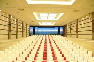 Sala de conferências com fileiras de cadeiras, uma janela ampla com vista e teto com iluminação embutida.