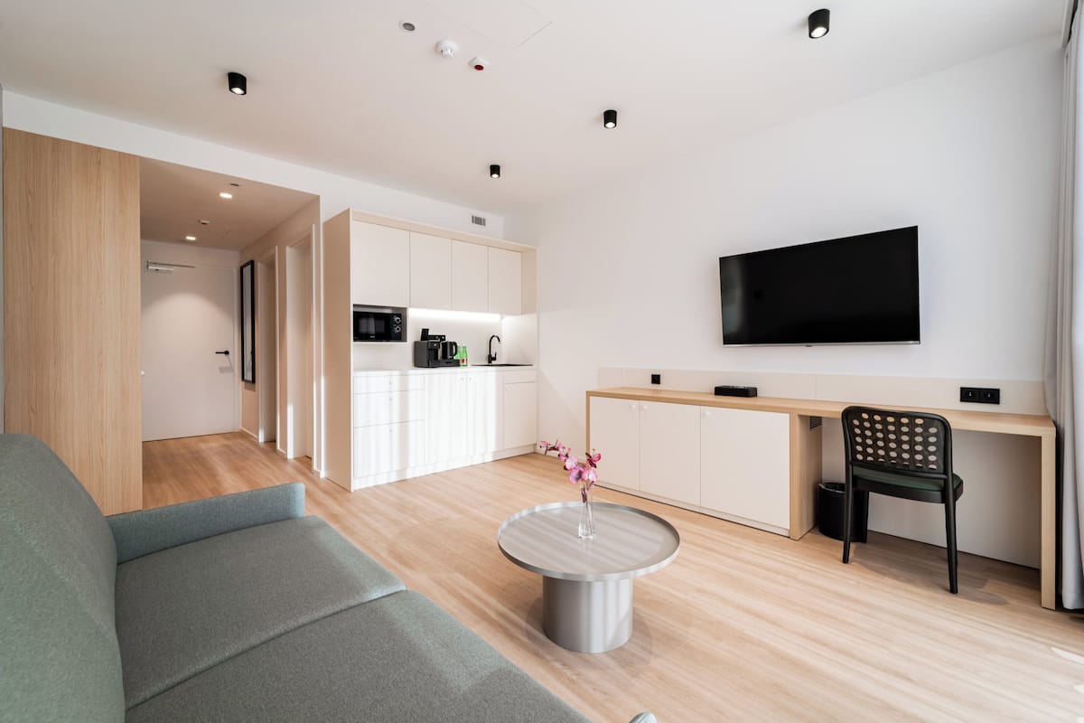 Sala de estar moderna com sofá cinza, mesa de centro redonda com um vaso, TV de tela plana na parede e área de cozinha ao fundo.
