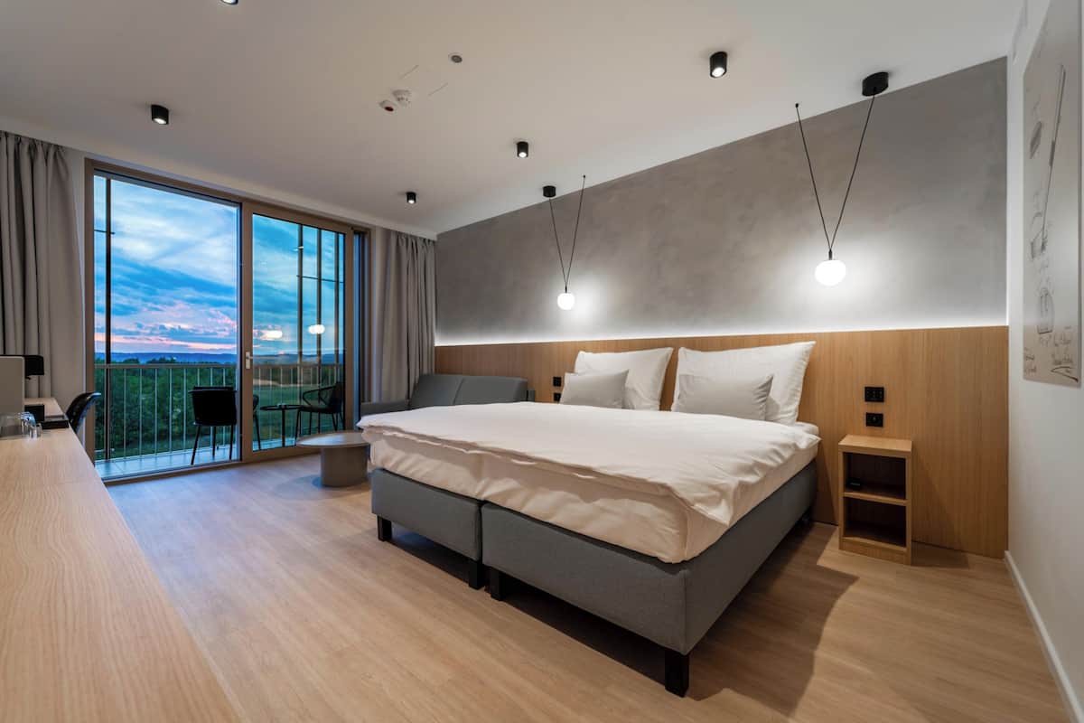 Quarto moderno com uma cama grande, uma mesinha de cabeceira de madeira, uma escrivaninha e uma varanda com vista.