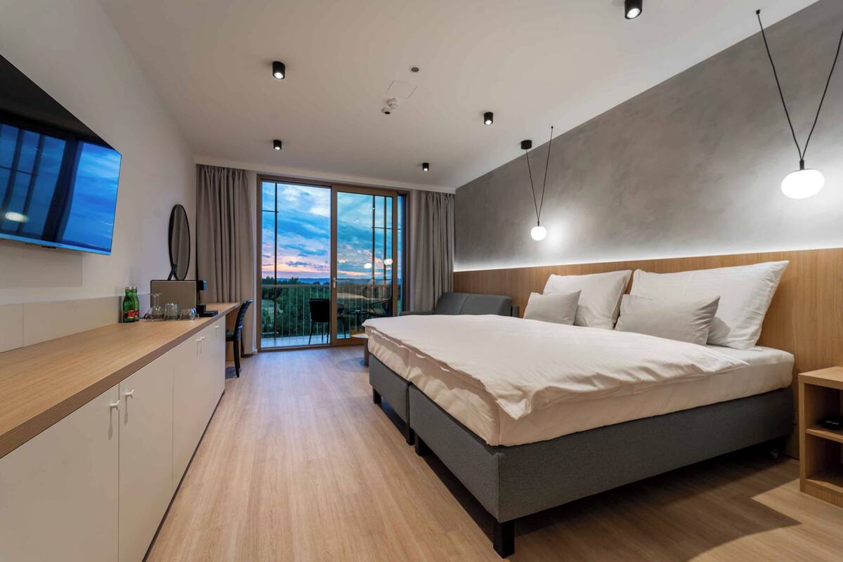 Quarto moderno com uma cama grande, televisão na parede, varanda com vista e escrivaninha de madeira.