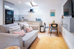 Uma sala de estar moderna com um sofá, uma mesa de jantar com cadeiras, uma cozinha com fogão e uma TV fixada na parede.