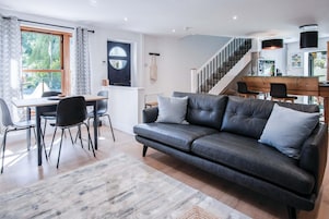 Uma sala de estar moderna com um sofá de couro escuro, uma área de jantar com mesa e cadeiras, uma escadaria e uma cozinha com balcão.