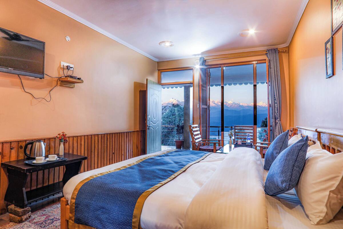 Quarto com cama, televisão na parede, mesinha com chaleira e chávenas, e vista para as montanhas através de uma porta aberta.