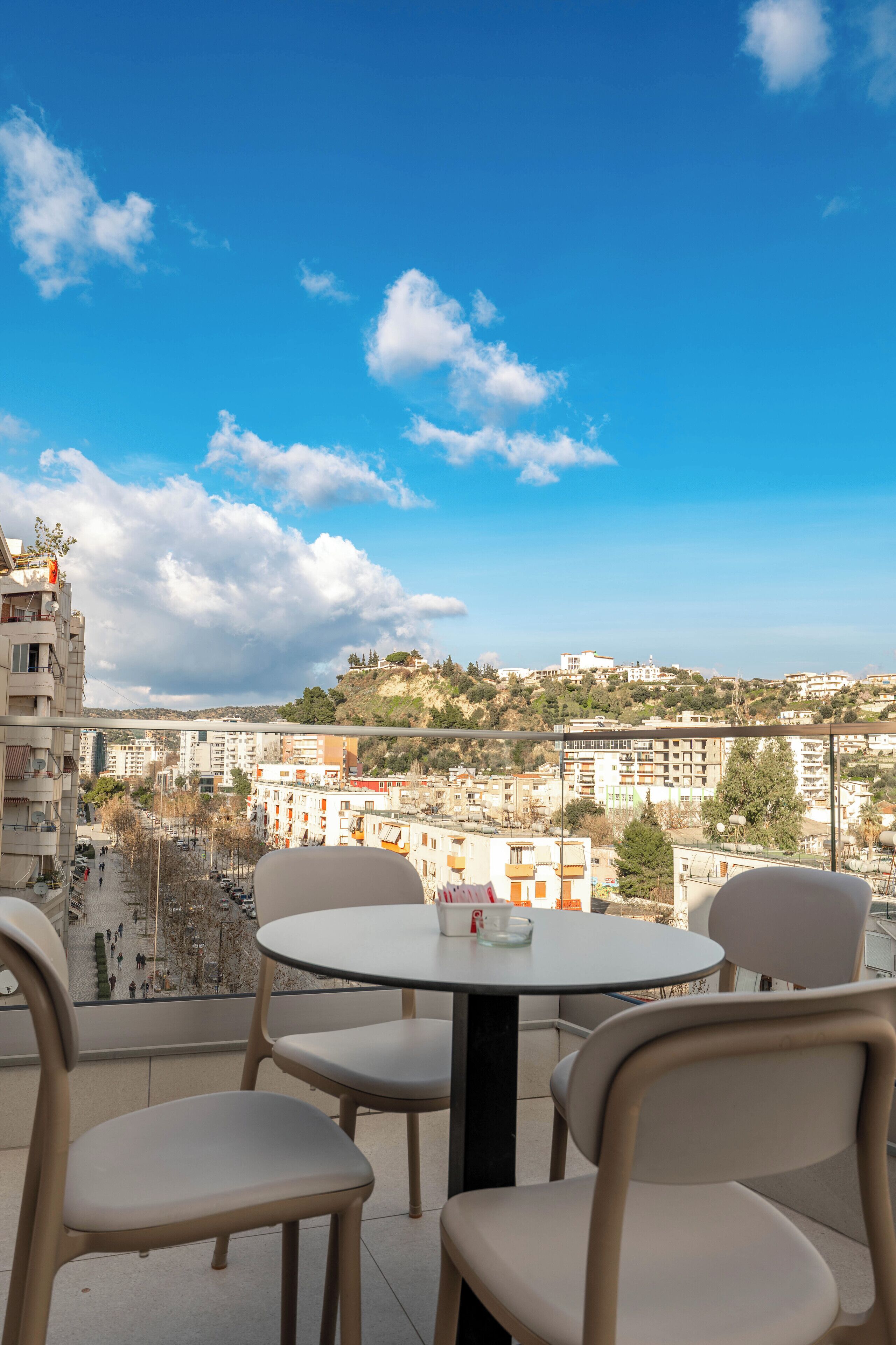 Uma varanda com uma mesa redonda e quatro cadeiras, com vista para uma paisagem urbana com edifícios e uma colina.