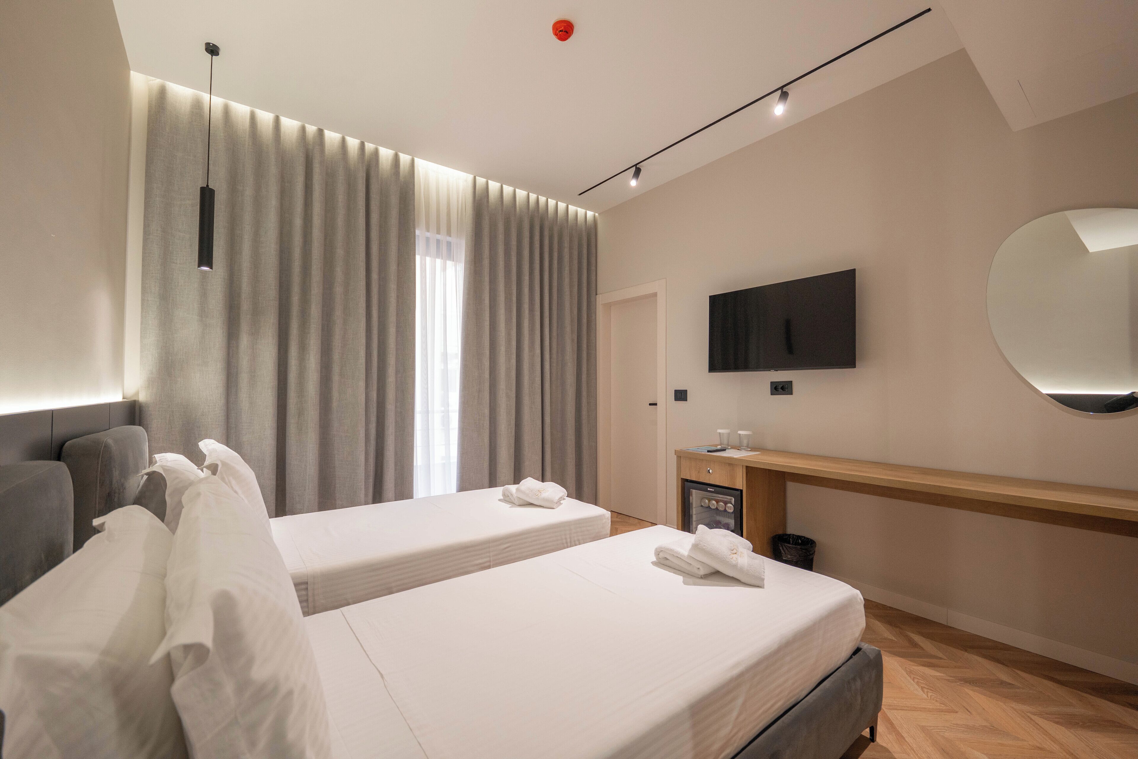 Quarto de hotel moderno com duas camas, televisão de tela plana, mesa de cabeceira de madeira e uma janela grande com cortinas.