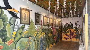 Um corredor com um mural de plantas e aves tropicais, quadros nas paredes e flores brancas pendentes.