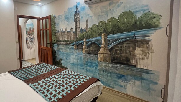 Quarto com um mural de uma ponte e torre do relógio, uma cama com roupa de cama estampada e um aparelho de ar condicionado na parede.