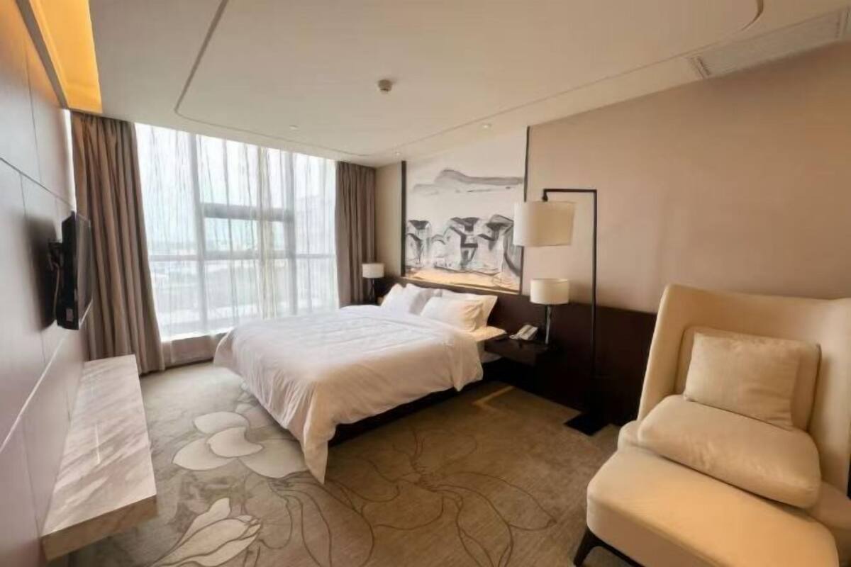 Quarto de hotel com uma cama, uma cadeira, uma escrivaninha, uma televisão e um quadro na parede.