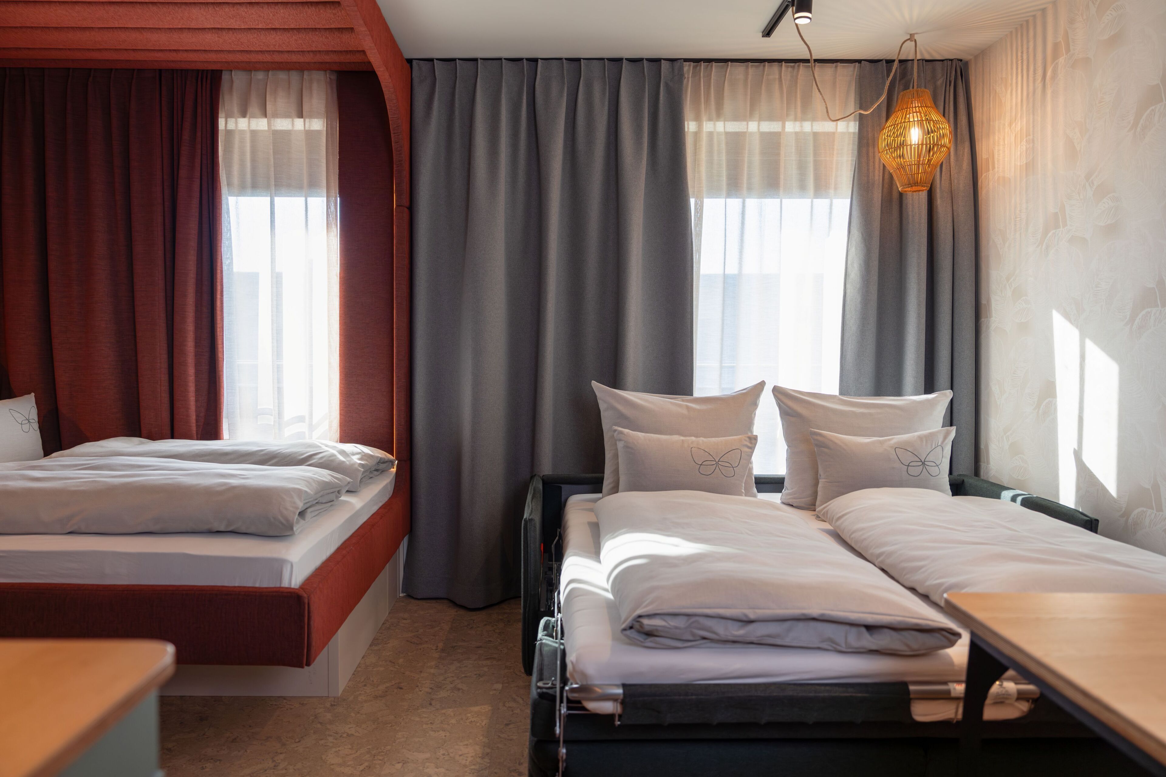 Quarto de hotel com duas camas, uma mesa de madeira, uma mala e um candeeiro suspenso.