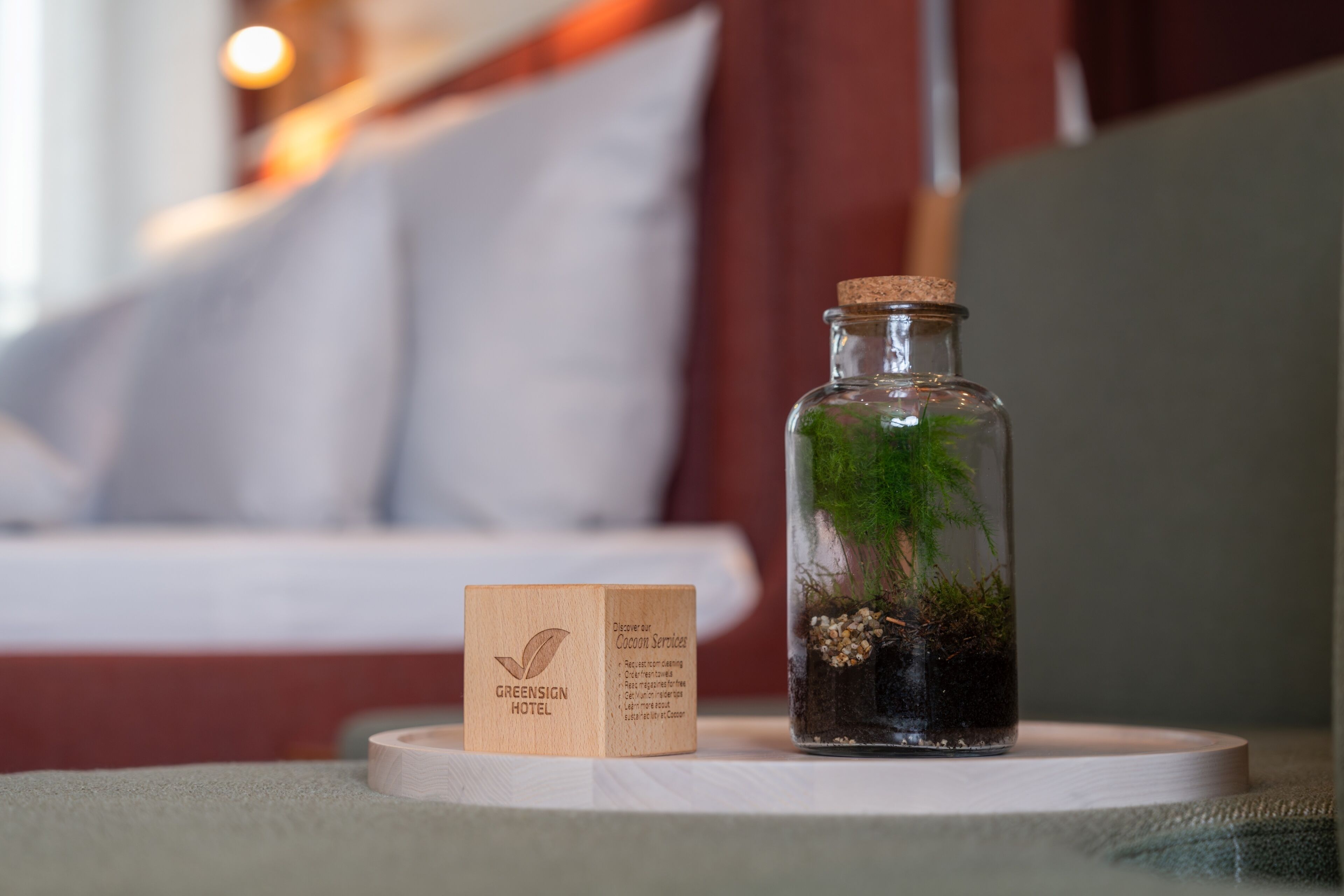 Uma garrafa de vidro com um rolhão de cortiça contendo uma pequena planta, colocada sobre uma superfície branca com um bloco de madeira com a inscrição 'Greensign Hotel' ao lado.