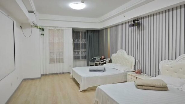 Quarto de hotel com duas camas, uma janela com cortinas, cabeceira branca, mesa de cabeceira com abajur e projetor fixado na parede.