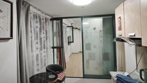 Quarto compacto com porta de vidro deslizante para um banheiro, uma pequena mesa com cadeira e um espaço com cozinha compacta.