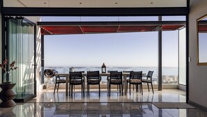Uma zona de refeições moderna com vista para o oceano, com uma mesa, cadeiras, uma churrasqueira e uma porta de vidro que dá para a varanda.