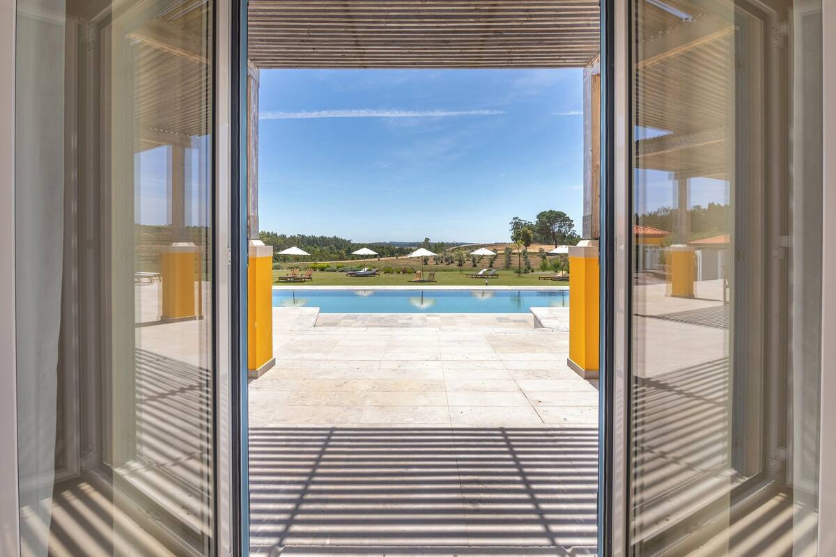 Vista através de uma porta de vidro para uma área de piscina, com céu claro, mobiliário externo e um jardim paisagístico.
