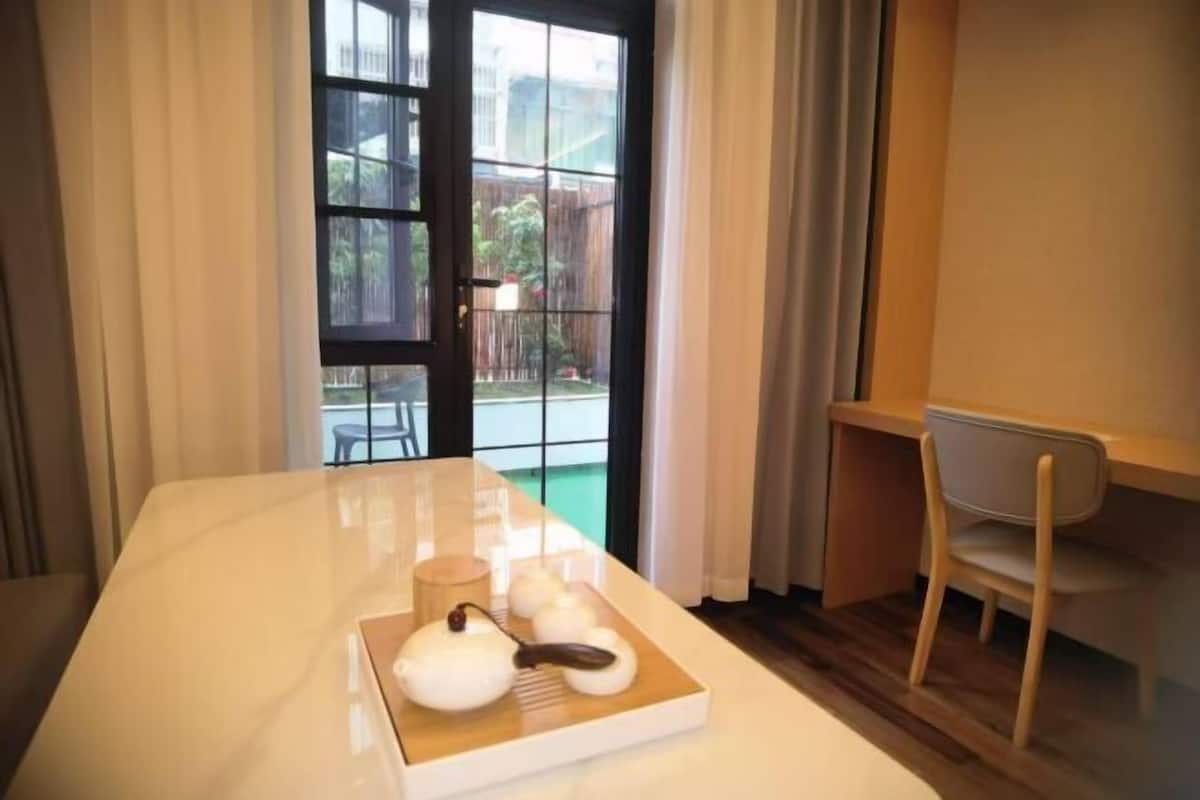 Quarto com mesa de madeira, uma cadeira, porta com vista para um jardim e janela com cortinas.