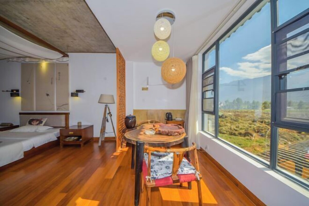 Quarto de hotel moderno com chão de madeira, uma cama, uma mesa de jantar com vista e grandes janelas.
