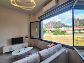 Quarto moderno com uma janela grande, TV de tela plana, sofá branco e uma mesa pequena.
