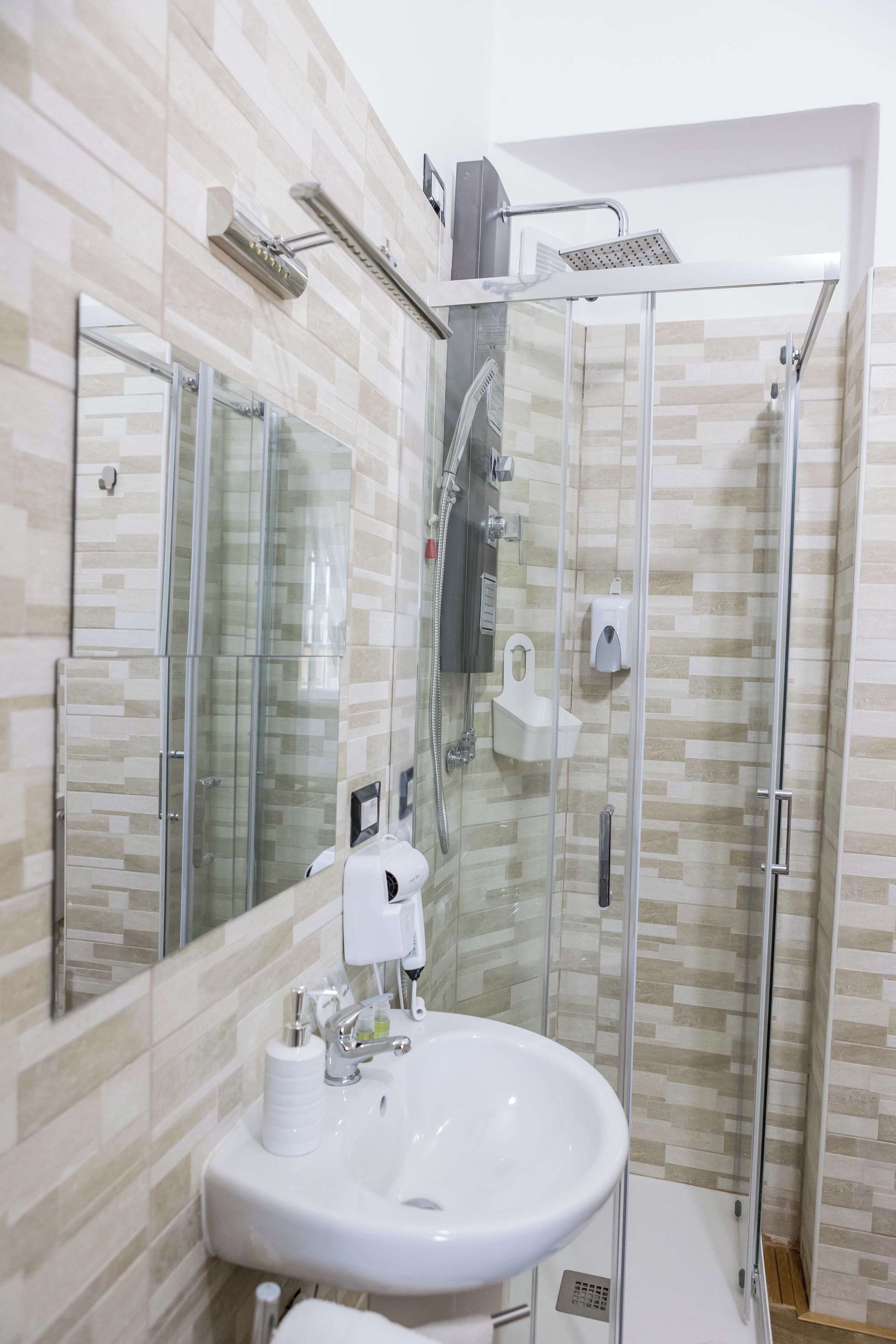 Casa de banho moderna com chuveiro em cabine de vidro, lavatório branco com espelho e azulejos bege estampados.