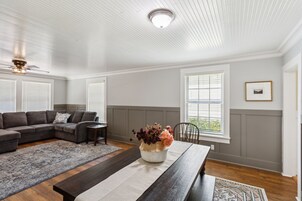 Uma sala de estar com sofá seccional, mesa de centro com vaso, mesa de jantar e ventilador de teto.