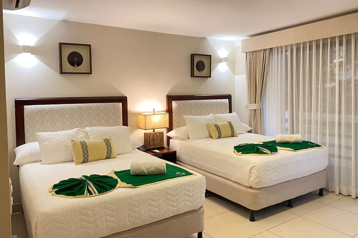 Quarto de hotel com duas camas, cada uma com roupa de cama branca e detalhes verdes, mesa de cabeceira com abajur e uma janela grande com cortinas.