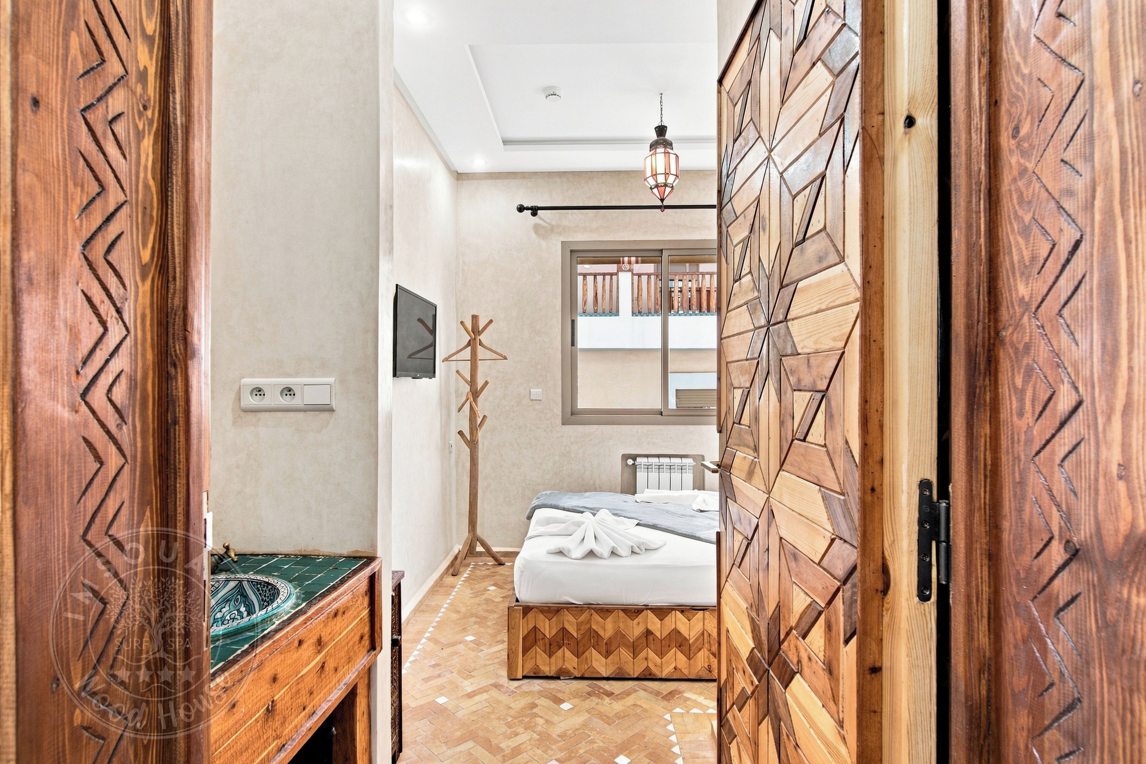 Quarto com piso em padrão herringbone, cama com roupa de cama branca, porta-roupas de madeira e luminária no teto.