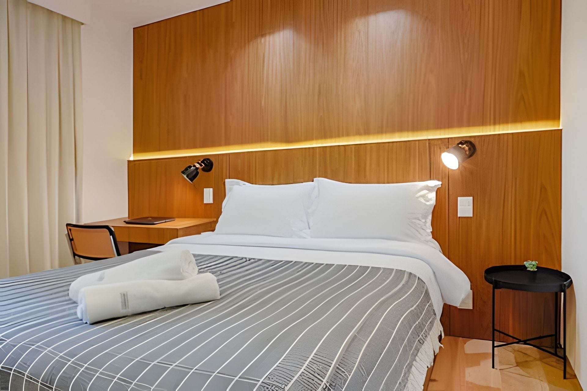Quarto de hotel com uma cama grande, uma mesa e duas luminárias fixadas na parede.