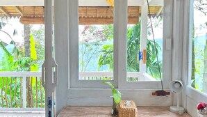 Quarto com escrivaninha de madeira, um banquinho, um ventilador e janela com vista para o verde.