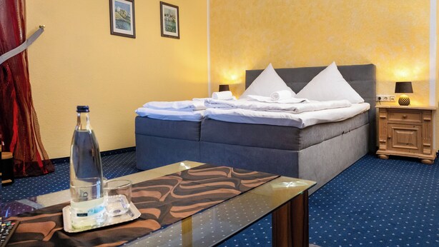 Quarto de hotel com cama, mesas de cabeceira, uma mesinha com garrafa e copos, e um tapete azul.