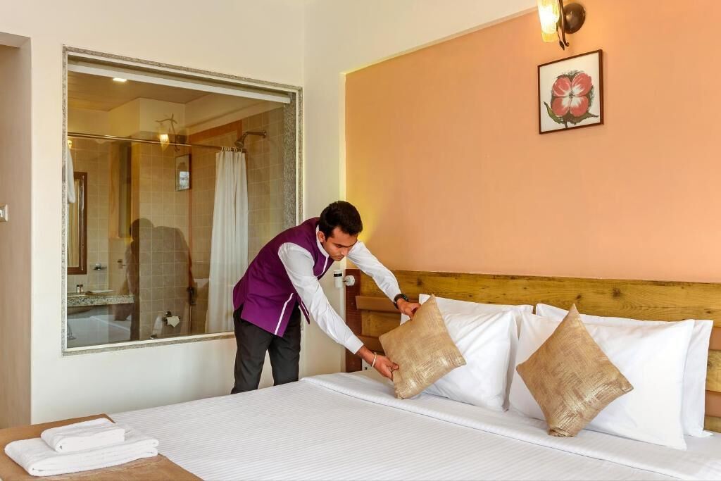 Quarto de hotel com cama, cabeceira de madeira, um espelho grande e banheiro visível através da porta de vidro.