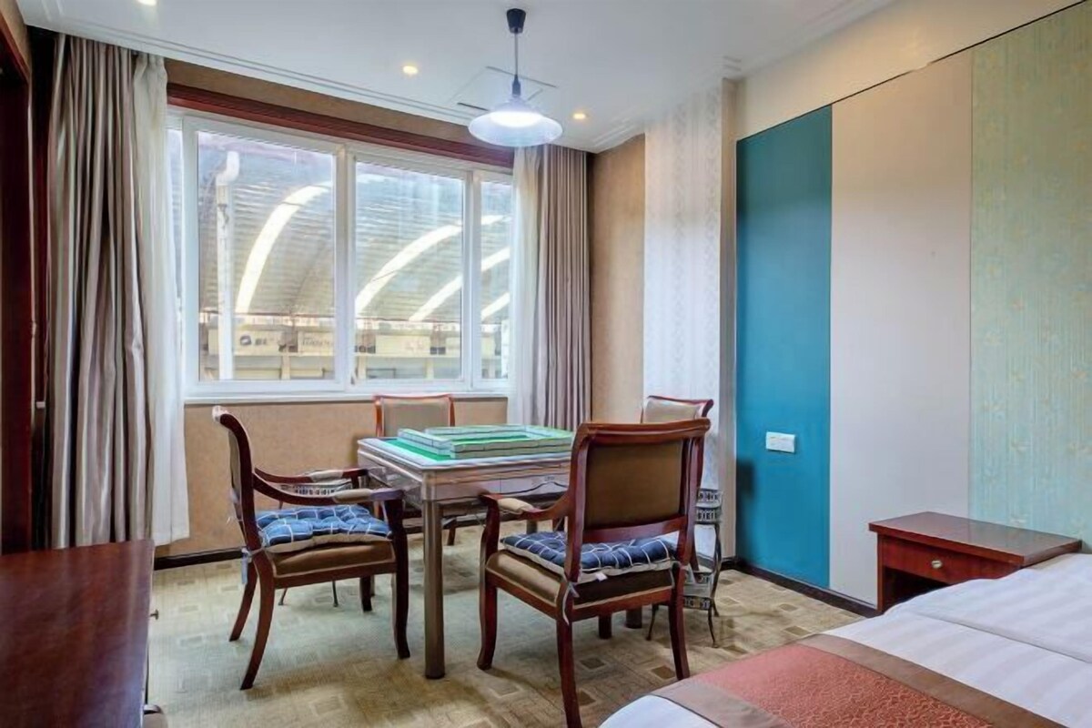 Quarto de hotel com uma cama, uma escrivaninha com cadeiras, uma janela com cortinas e vista para um edifício.